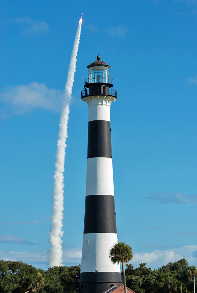 A rocket launch streaks skyward beside the historic lighthouse, capturing the perfect marriage of 19th-century maritime safety and 21st-century space exploration.