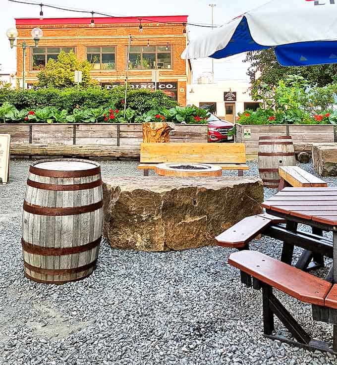The outdoor seating area offers rustic charm with barrel tables, stone fire pit, and the perfect atmosphere for summer barbecue sessions.