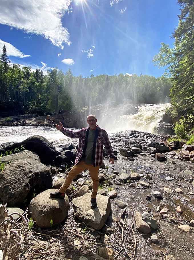 A delighted explorer strikes a pose before the roaring waterfall, capturing that perfect "I conquered the trail" moment at journey's end.