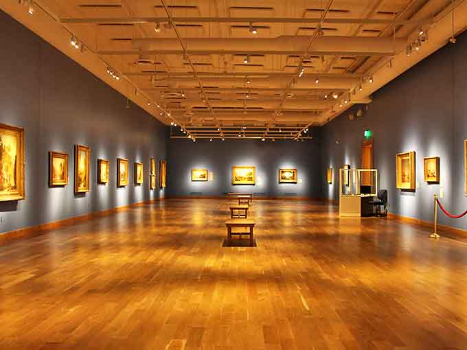 Interior: Sunlight dances across polished hardwood floors in this gallery, where masterpieces hang against deep blue walls like treasures from a sophisticated shipwreck.