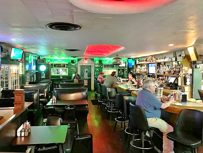 Interior: Green neon casts a welcoming glow across the bar where regulars gather and newcomers become friends. Classic dive bar magic.