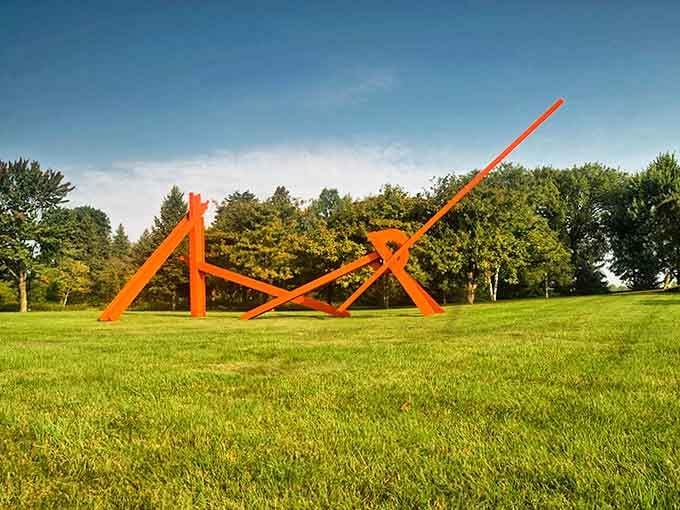 Fiery orange beams reach skyward like a modernist matchstick construction, casting dramatic shadows that become part of the artwork itself.