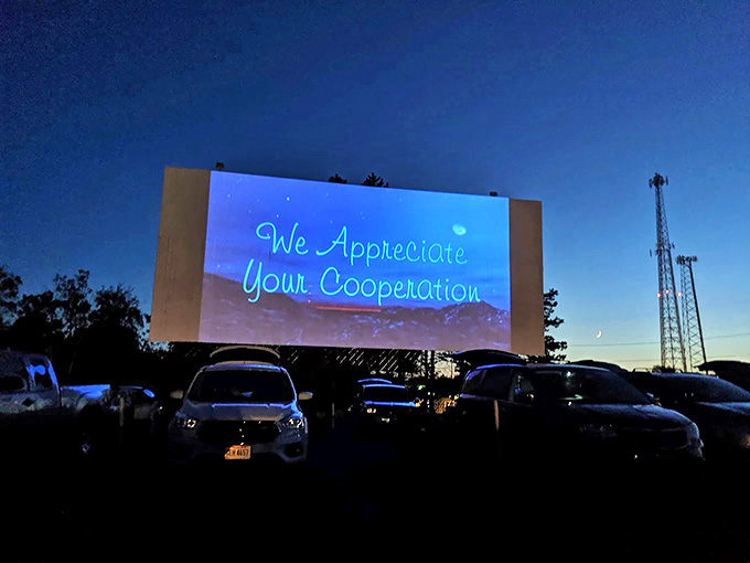 We Appreciate Your Cooperation glows in neon blue against the twilight sky – a gentle reminder of drive-in etiquette as darkness falls.