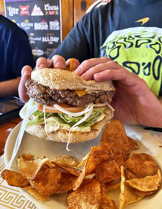 Hands cradle this magnificent burger creation like it's precious cargo. Look at those layers! That's not dinner, that's architecture with cheese.