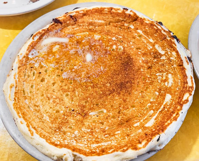 A perfect buttermilk pancake in its natural habitat – golden-brown, plate-sized, with butter melting into a heavenly puddle at its center.
