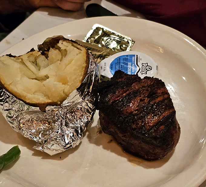 A perfectly charred filet mignon alongside a foil-wrapped baked potato &ndash; simplicity elevated to art form on a white plate.