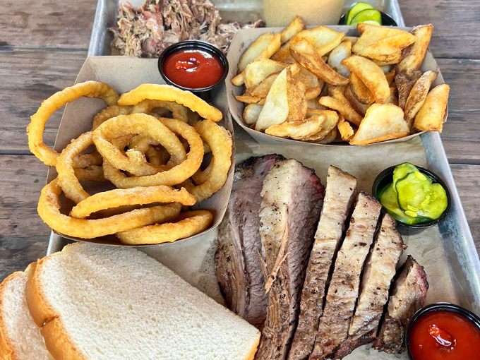 A barbecue feast fit for royalty: tender sliced brisket, golden onion rings, and house-cut potato wedges served unpretentiously on metal trays.