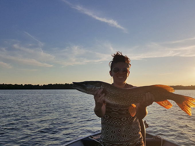 A proud angler displays her impressive northern pike catch against Deer Lake's golden sunset &ndash; the perfect Minnesota fishing moment.