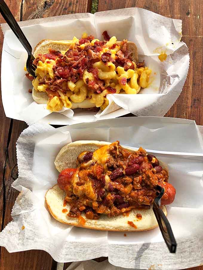 Two hot dog masterpieces: one crowned with bacon-studded mac & cheese, the other a hearty chili creation &ndash; proof that simplicity and indulgence aren't mutually exclusive.