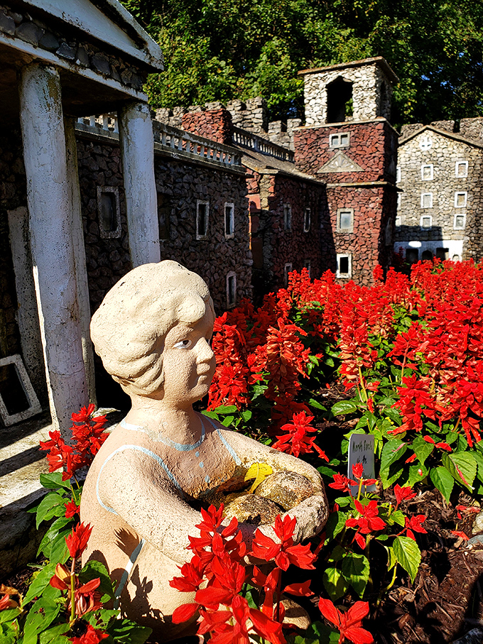 A serene garden statue stands sentinel among vibrant red flowers, creating a peaceful moment amid the garden's more elaborate stone creations.