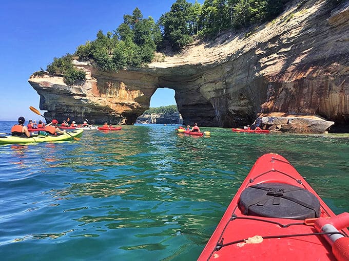 Kayakers paddle beneath towering cliffs toward natural stone arches, gaining perspectives of Pictured Rocks impossible to experience any other way.