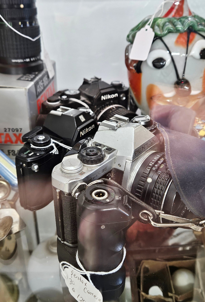 These vintage Nikon and Pentax cameras once captured family vacations and first steps, mechanical marvels from when "developing" meant chemicals, not clicking an app icon.