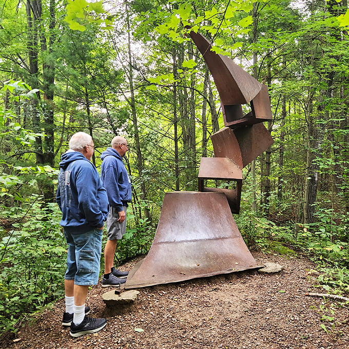 Two visitors contemplate this towering rust-colored sculpture that seems to be reaching for the treetops with industrial determination.