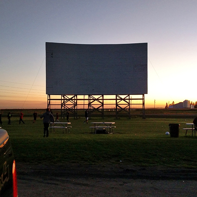 Silhouettes of moviegoers gather beneath the screen at dusk, creating human constellations against the fading light.