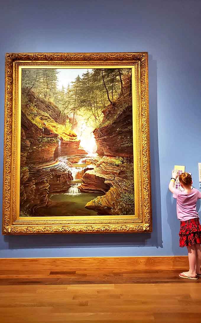 Massive painting: A young visitor stands transfixed before this luminous landscape, where sunlight breaks through forest canopy to illuminate cascading waters below.