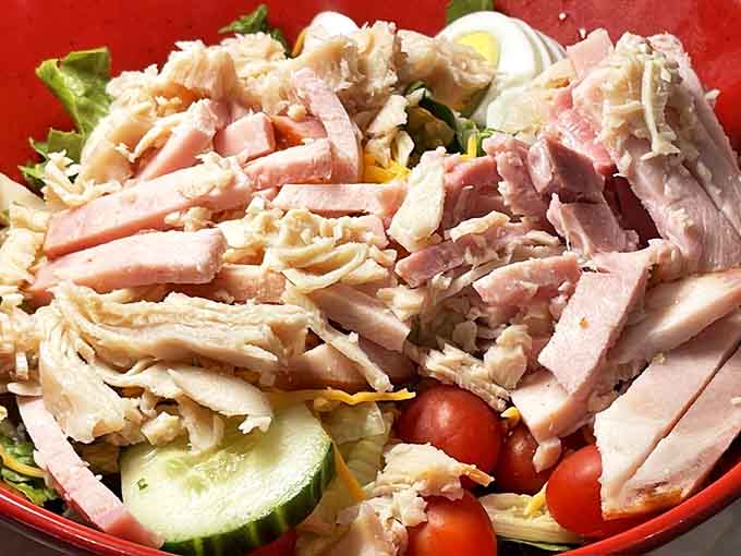 A mountain of freshness topped with protein confetti &ndash; julienned ham, turkey, and cheese creating a carnival of flavors atop crisp greens.