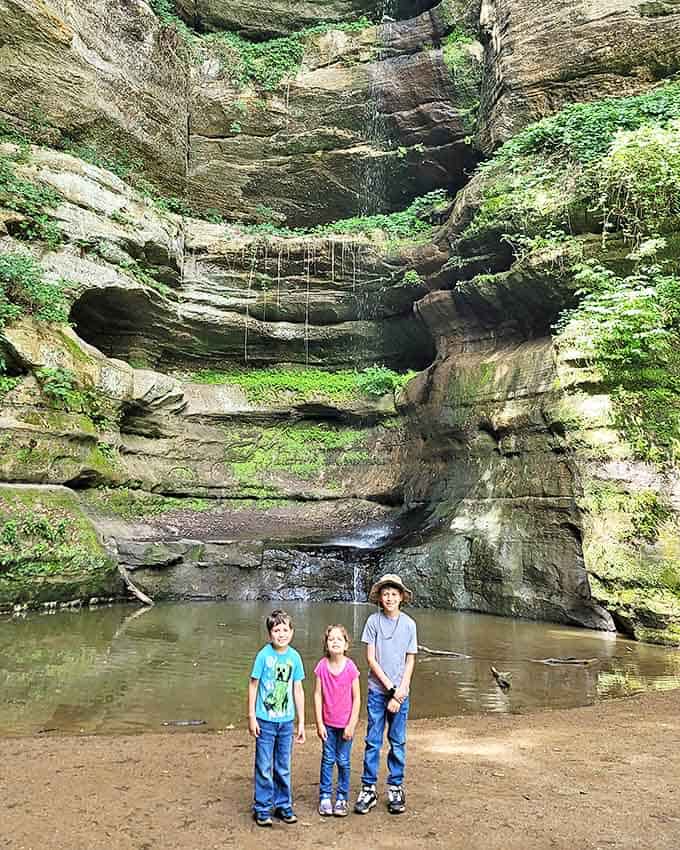 Three young adventurers pose before the majestic waterfall at Wildcat Canyon, their expressions capturing the wonder that awaits in Starved Rock's most dramatic gorge.