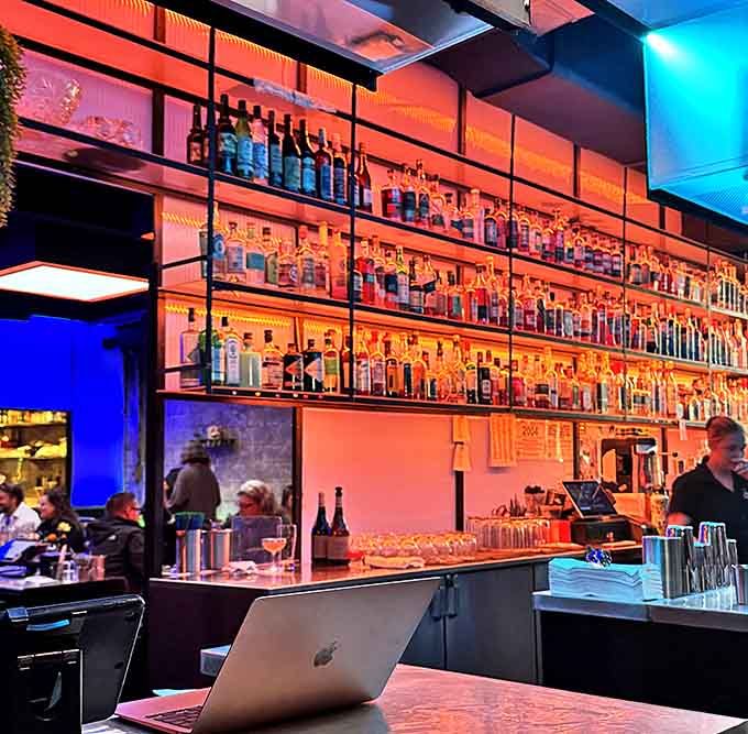 The bar shelves climb toward heaven, bottles backlit like precious artifacts in this temple of food and drink.