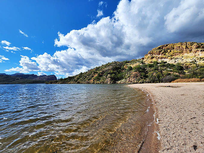 Butcher Jones Beach stretches along Saguaro Lake's shoreline, where golden sand meets crystal-clear water beneath dramatic desert cliffs.