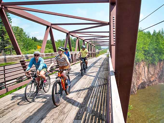 Iron-red bridge spans across the water as cyclists enjoy the Gitchi-Gami Trail, where wilderness meets engineering in perfect harmony.