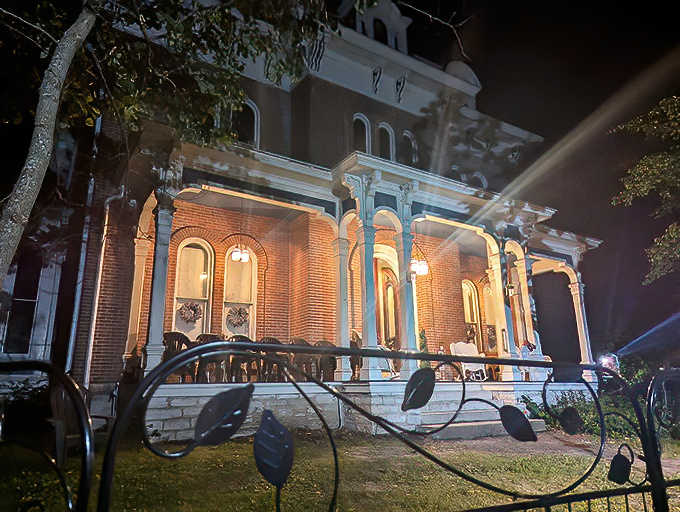 The mansion's illuminated facade creates an eerily beautiful tableau against the night sky, like a lighthouse for wandering spirits.