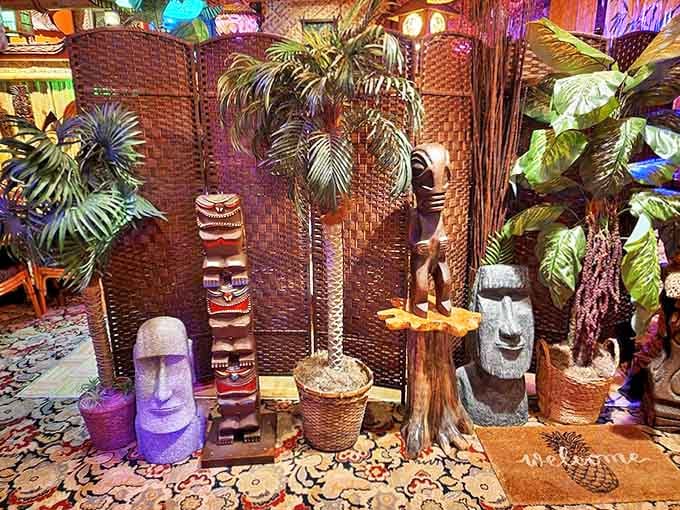 Tiki statues stand guard among lush tropical plants and woven screens. These wooden sentinels create distinct dining areas while maintaining the immersive Polynesian atmosphere.