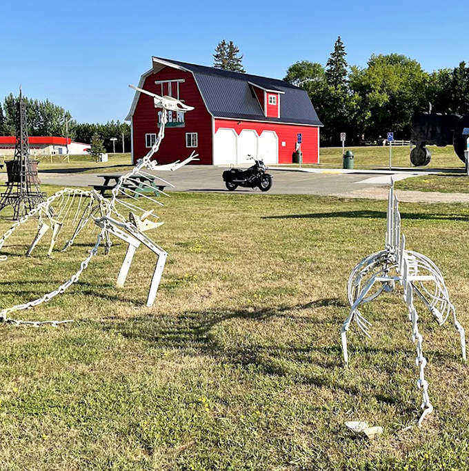 White skeletal sculptures create an otherworldly forest, their bone-like structures standing in stark contrast to the vibrant red barn.