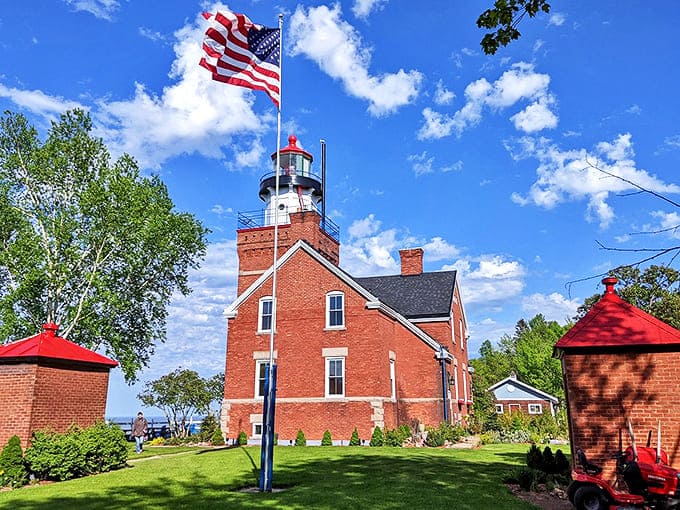 American pride flies high above the historic lighthouse, its red brick tower and white lantern room standing tall against a perfect blue sky.