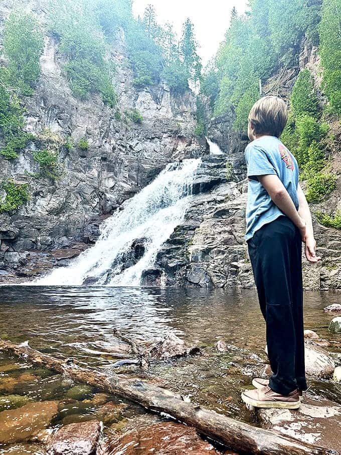 A young observer contemplates the waterfall's hypnotic rhythm from a perfect vantage point. Some lessons can only be taught by nature itself.