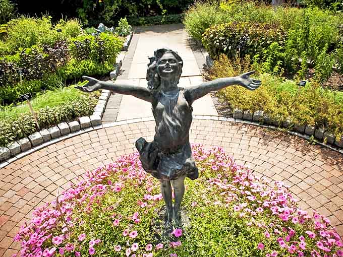 This bronze girl with arms outstretched captures pure joy, surrounded by pink flowers as if celebrating the simple pleasure of sunshine.