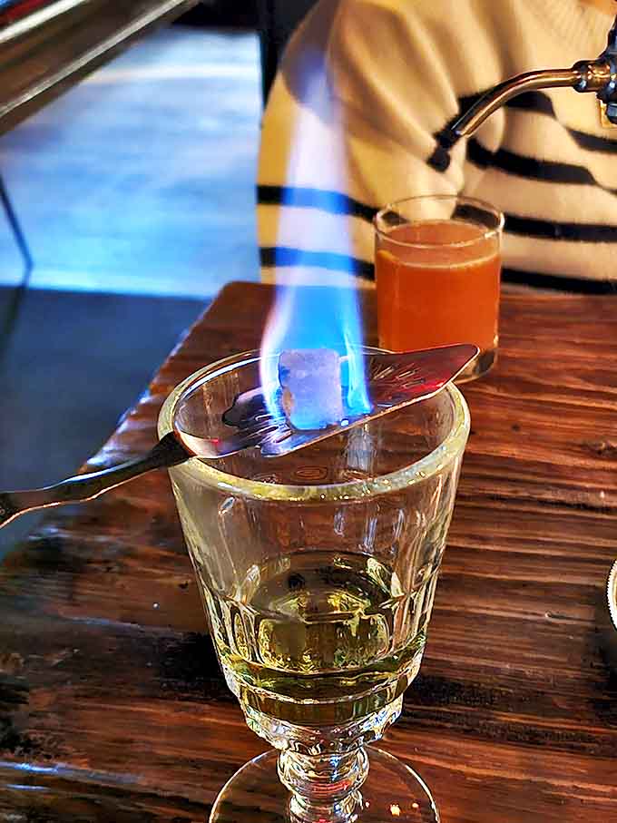 The absinthe fountain transforms drinking into ceremony, as water slowly drips over sugar cubes, releasing essential oils in a hypnotic cloudy swirl.