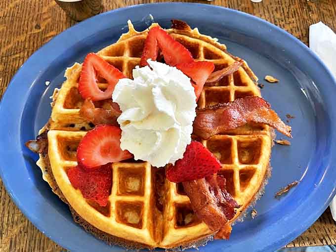 Breakfast nirvana achieved: A golden waffle crowned with fresh strawberries, whipped cream, and bacon that's practically begging to be photographed before it's devoured.