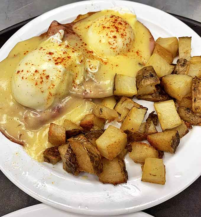 Eggs Benedict draped in hollandaise gold atop Canadian bacon, with home fries providing crispy dividends on the side of perfection.