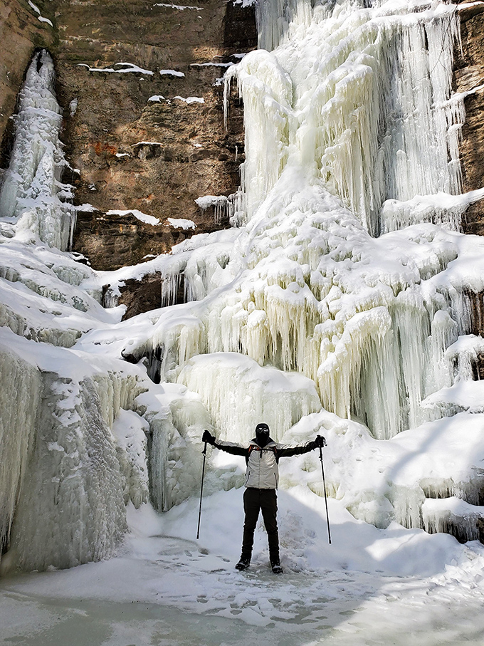 A solitary explorer stands triumphant before a frozen waterfall, arms outstretched as if embracing winter's spectacular ice sculpture.