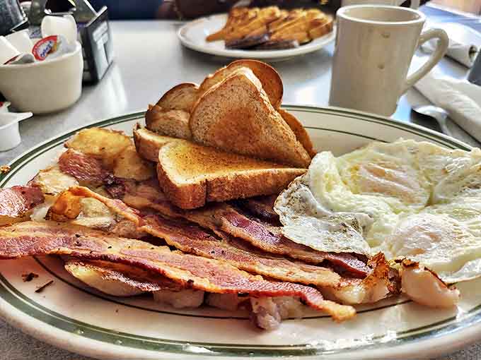 Breakfast perfection on a plate – crispy bacon arranged like fallen dominoes beside sunny eggs and toast waiting for its butter bath.