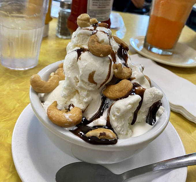 Hot fudge cascades over vanilla scoops like a chocolate waterfall, with cashews standing guard against too-quick consumption.