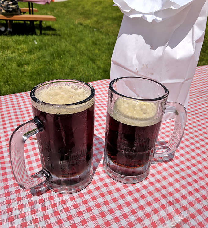 Two frosty mugs of homemade root beer on a classic red checkered tablecloth &ndash; a scene that could be from yesterday or fifty years ago.