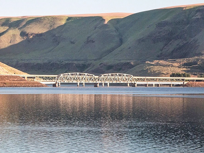 The train crosses a steel bridge spanning calm waters, reflecting the surrounding hills in perfect symmetry.