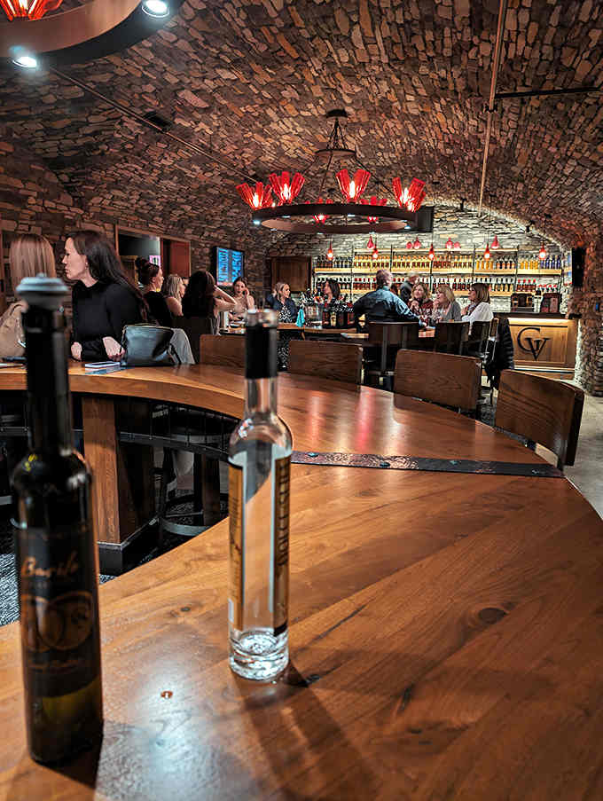 Stone walls curve overhead like a wine cellar from medieval times, while modern patrons gather at wooden tables, clinking glasses beneath dramatic red lighting.