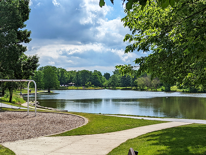 Patriots Park's serene pond reflects clouds drifting lazily overhead, while nearby benches invite visitors to pause and appreciate the view.