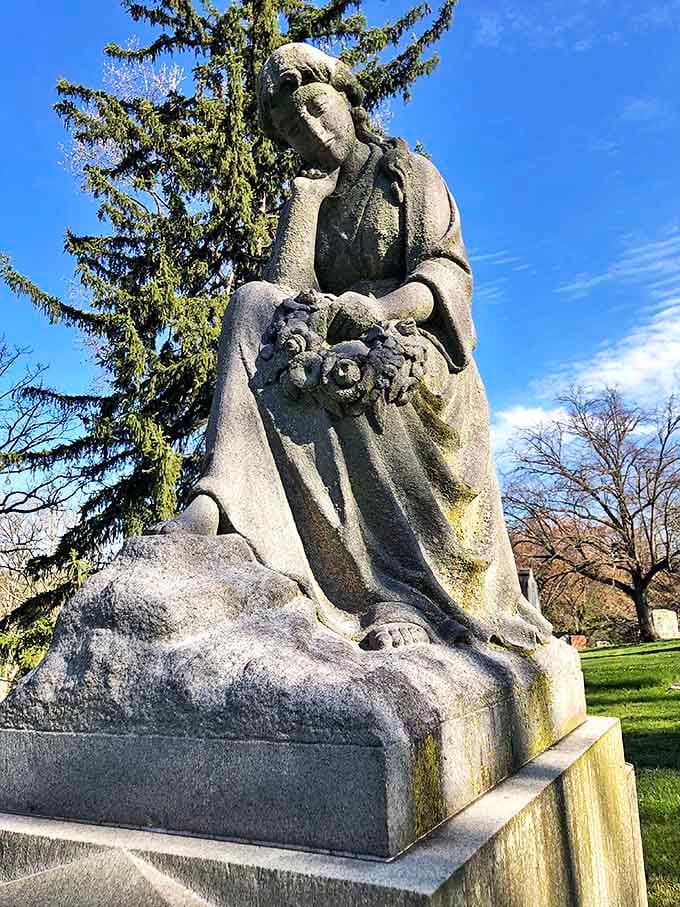 A pensive stone figure cradles flowers, her weathered face showing the gentle patina that only decades of seasons can create.