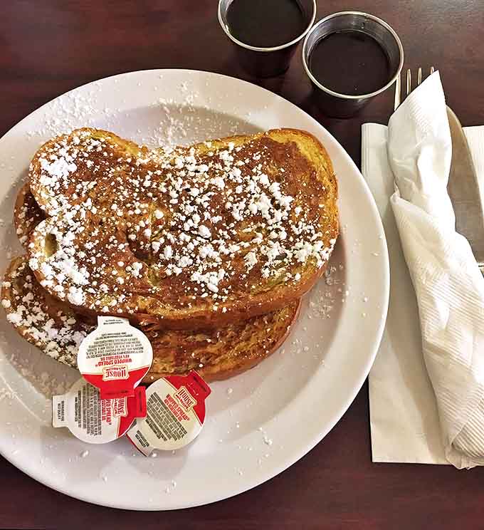 Thick-cut French toast achieves golden perfection with powdered sugar snow, accompanied by syrup and butter for maximum morning indulgence and happiness.