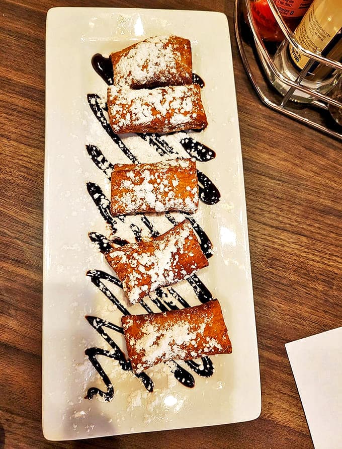 Pillowy squares of fried dough wearing powdered sugar like freshly fallen snow, with chocolate drizzles creating delicious abstract art.