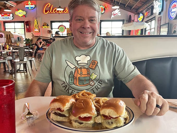 A smiling patron enjoys sliders served on classic diner plates, surrounded by the warm glow of neon signs&mdash;proof that happiness can be found between two buns.