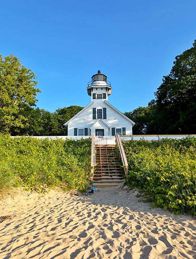 The lighthouse beckons visitors up sandy steps, its white picket fence and green surroundings creating a quintessentially American maritime scene.