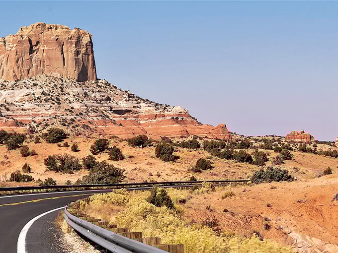 A ribbon of black asphalt winds through crimson desert, with towering mesas standing guard against an impossibly blue Arizona sky.