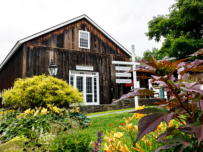 The weathered wooden exterior of Phelps Barn welcomes visitors with rustic charm, colorful gardens, and the promise of Vermont hospitality inside.