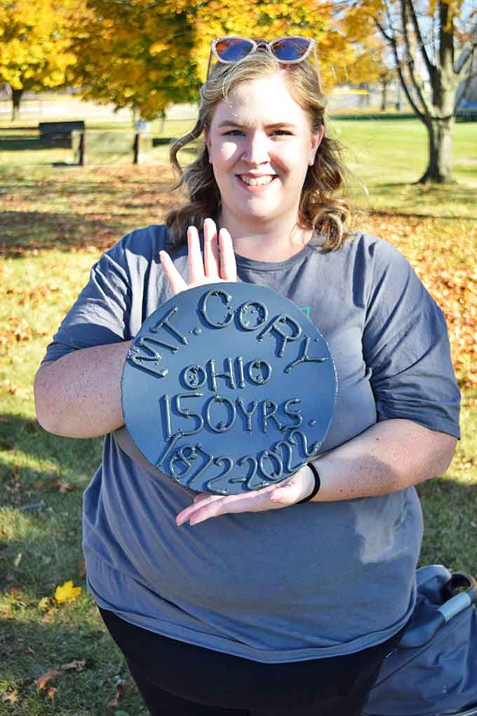 A proud resident displays the commemorative plaque celebrating Mount Cory's 150th anniversary &ndash; a milestone marked by community rather than commerce.