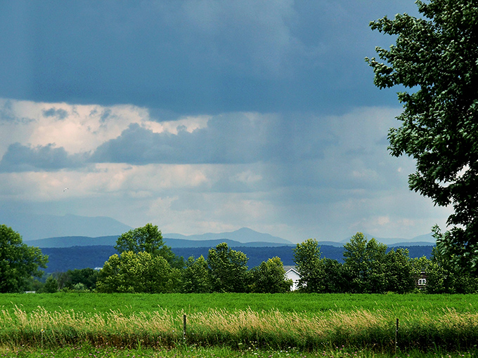 Storm clouds gather dramatically over Grand Isle's countryside, painting the landscape in moody blues and emerald greens.
