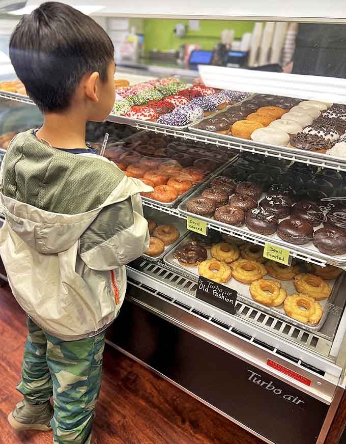 A young customer contemplates the wall of possibilities, his expression capturing that universal moment of donut-selection deliberation we all understand.